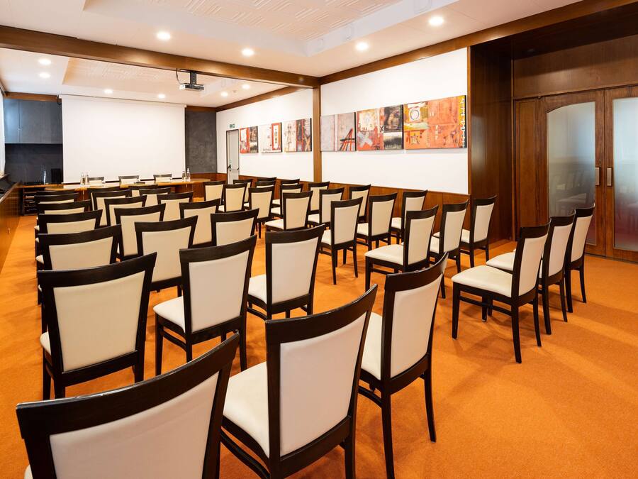 Uma sala de conferências com fileiras de cadeiras, uma tela de projetor e obras de arte nas paredes.