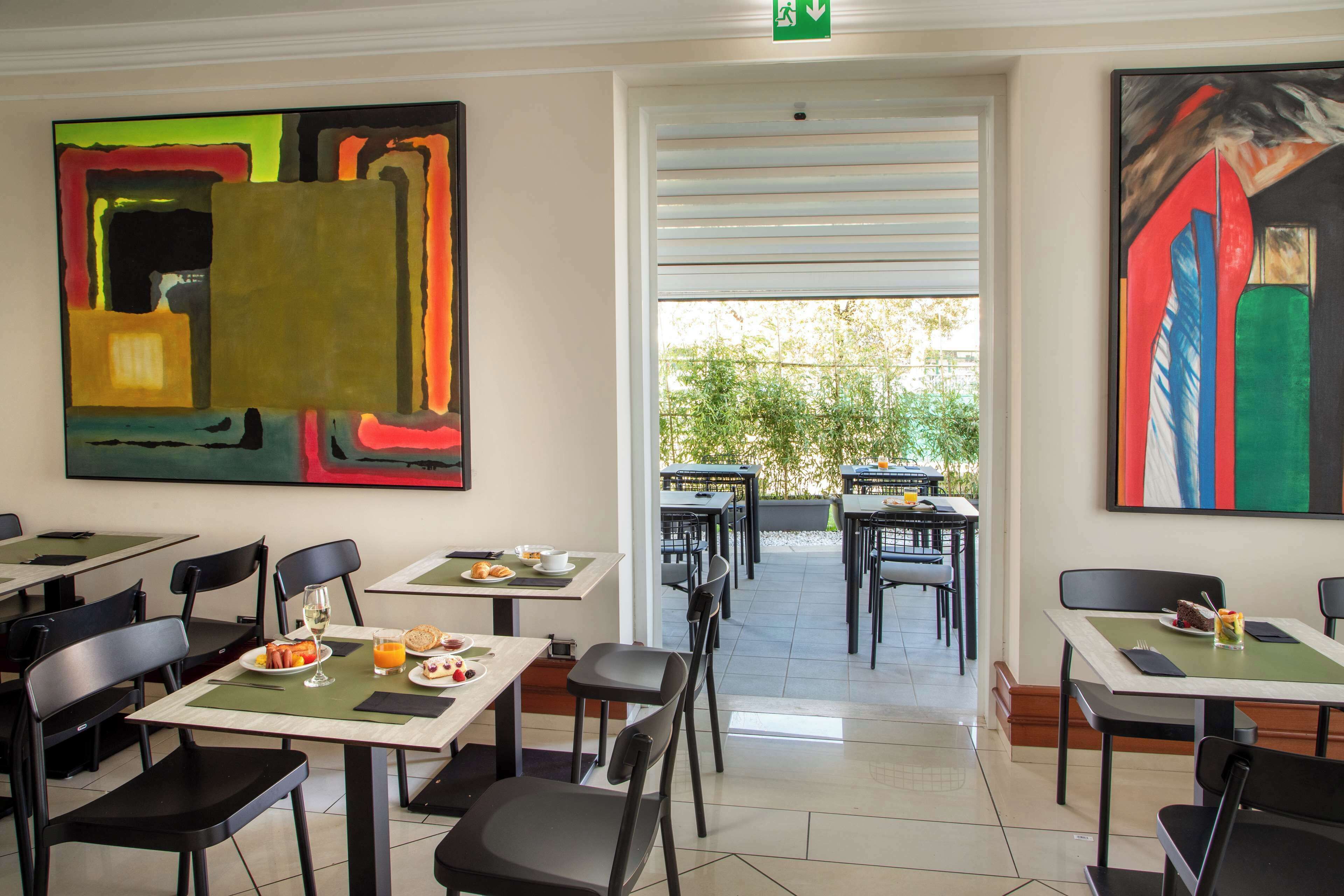 Uma sala de jantar moderna com mesas e cadeiras, pinturas abstratas grandes nas paredes e vista para o verde lá fora.