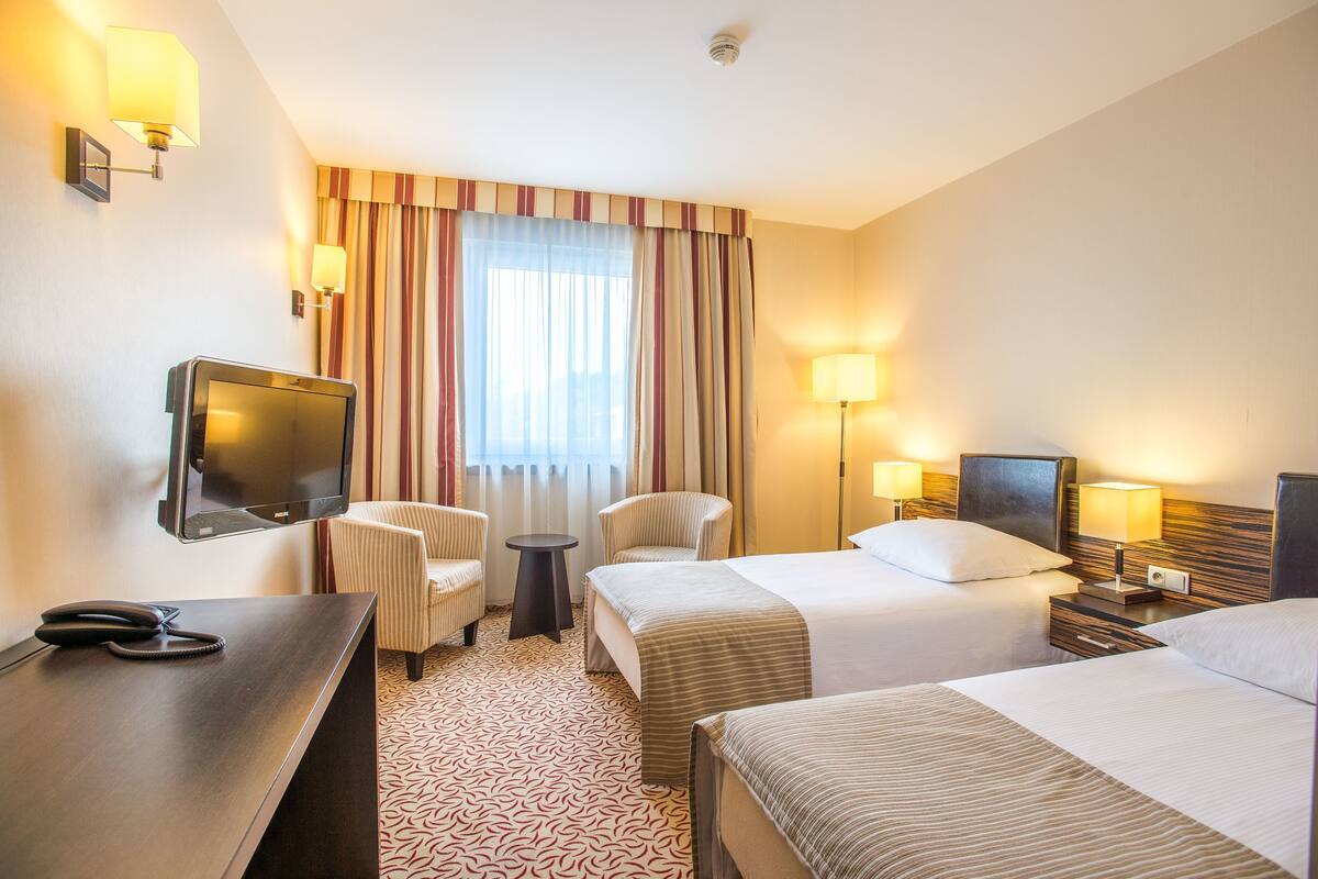 Quarto de hotel com duas camas, televisão, escrivaninha com telefone e duas cadeiras.