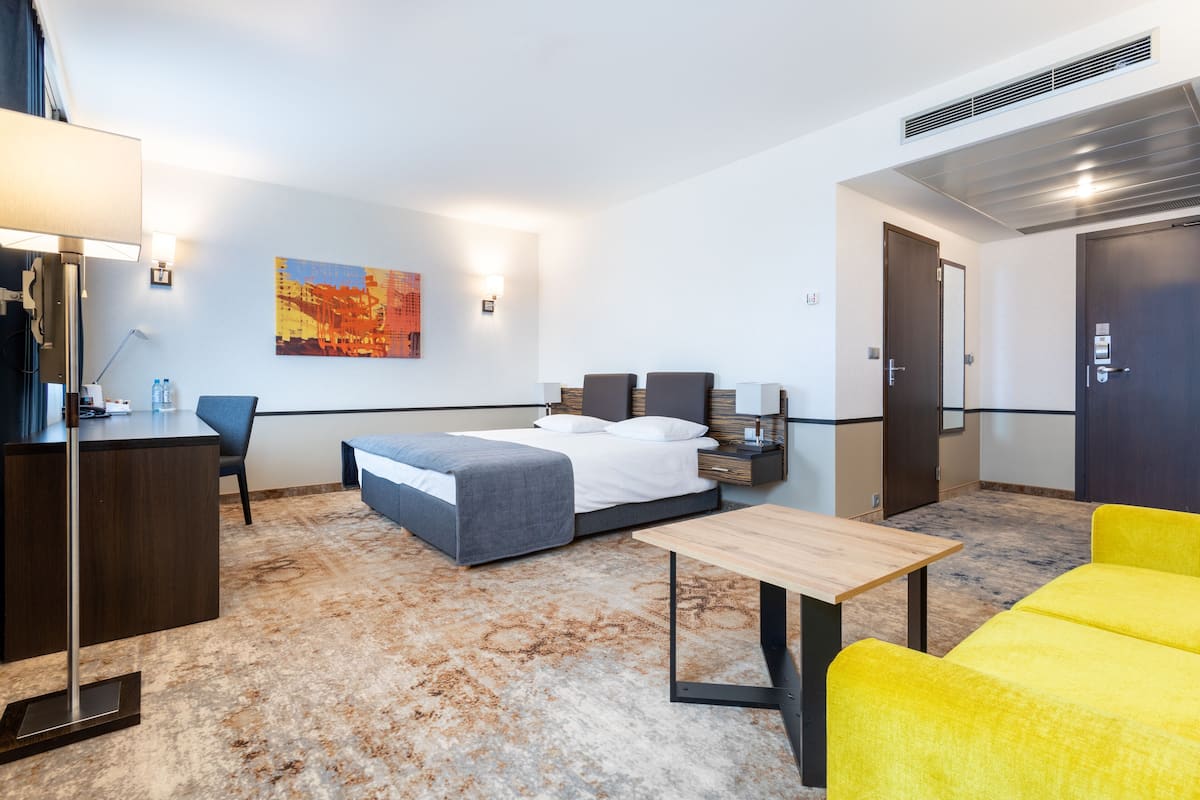 Quarto de hotel com cama, mesa de cabeceira, escrivaninha, cadeira e um sofá amarelo vibrante.
