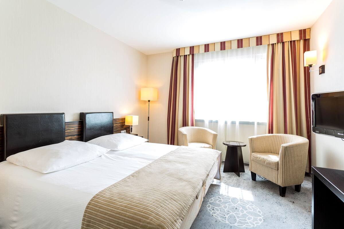Quarto de hotel com uma cama grande, duas mesas de cabeceira, uma cadeira, uma mesinha e uma televisão.