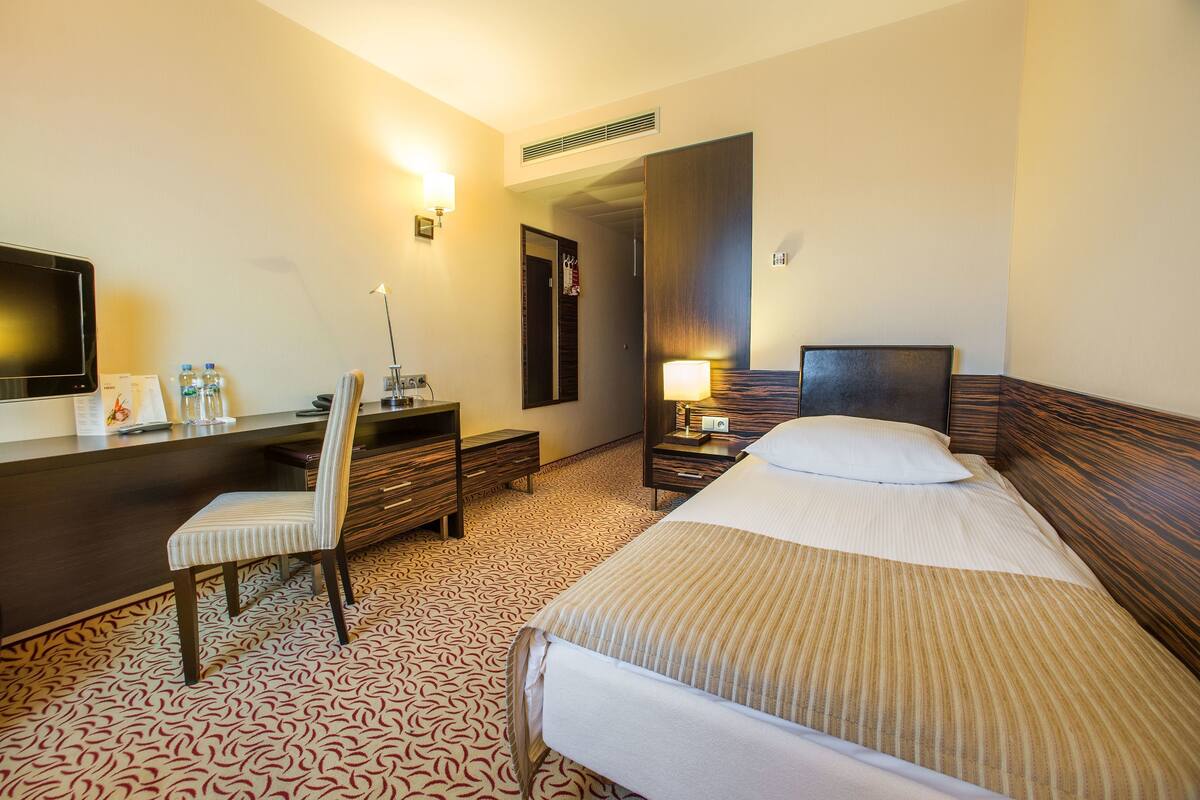 Quarto de hotel com cama, escrivaninha, cadeira, televisão e mesa de cabeceira com abajur.