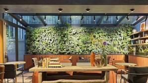 Uma área de refeições com uma mesa de madeira rústica, cadeiras e piso de azulejos. Uma parede verde grande com plantas fica atrás da mesa.