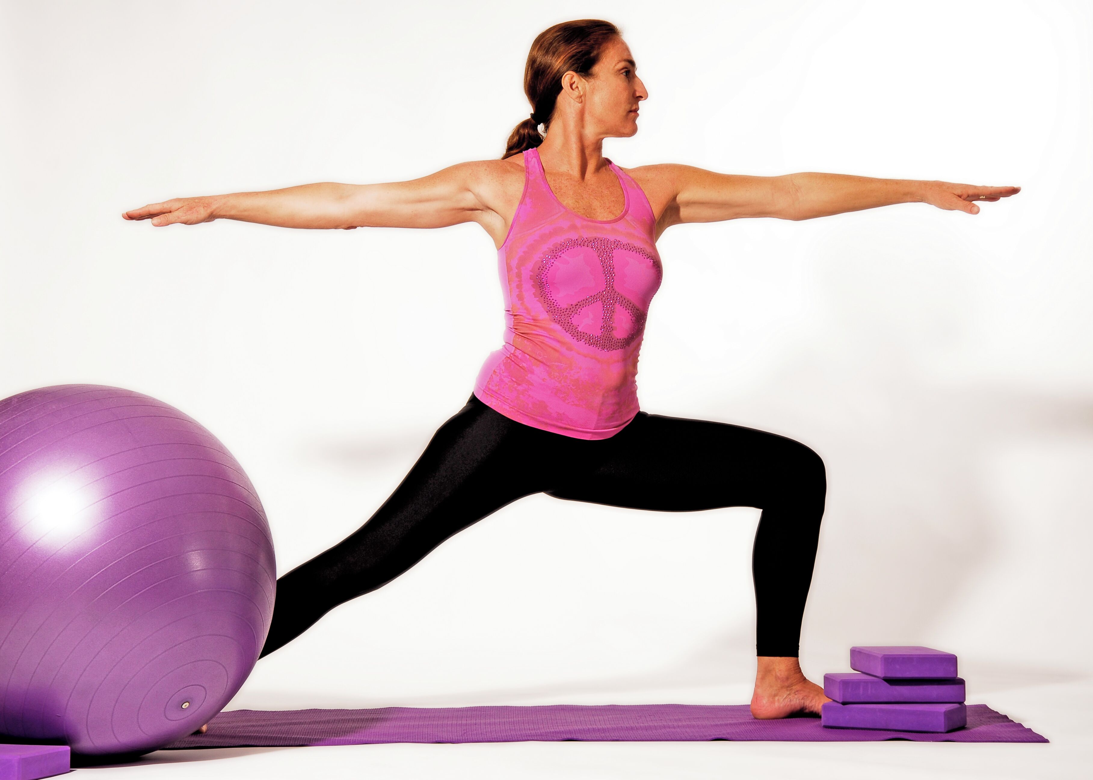 Uma pessoa fazendo uma pose de yoga em um tapete roxo, com uma grande bola de exercício roxa e uma pilha de blocos roxos.