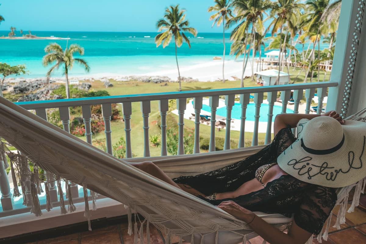 Uma pessoa relaxando em uma rede em uma varanda com vista para uma praia, palmeiras e uma piscina.