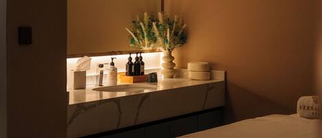 Quarto de hotel com cama, toucador com amenities, espelho e um vaso com flores.