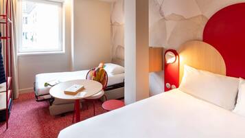 Quarto de hotel com duas camas, uma mesinha, uma cadeira, janela com cortinas e uma parede de destaque vermelha.