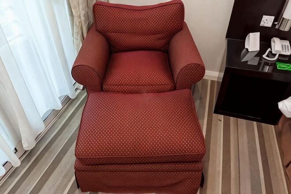 Uma poltrona vermelha estofada e um banquinho em um quarto com piso listrado, uma escrivaninha com telefone e uma janela com cortinas.