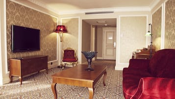Quarto de hotel com televisão, mesa de centro de madeira, sofá vermelho, carpete estampado e luminária decorativa.