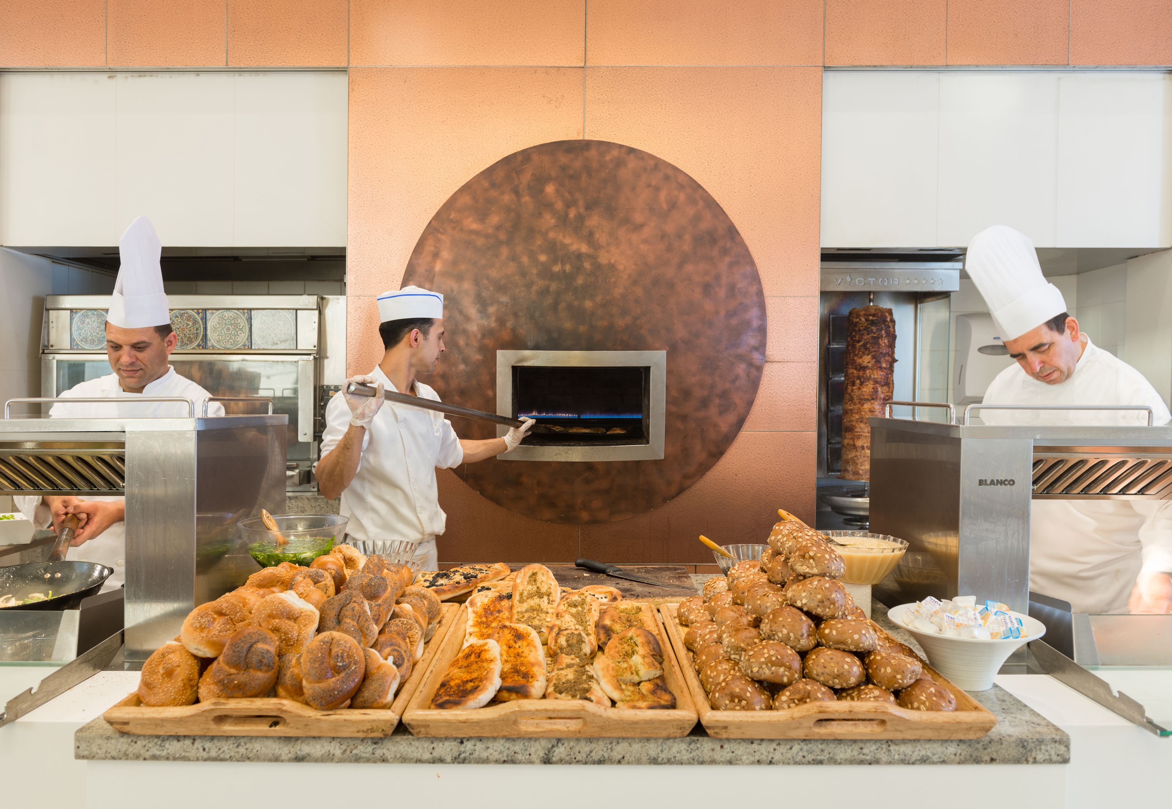 Uma cozinha comercial com chefs preparando alimentos, um forno grande e diversos produtos de panificação expostos em tábuas de madeira.