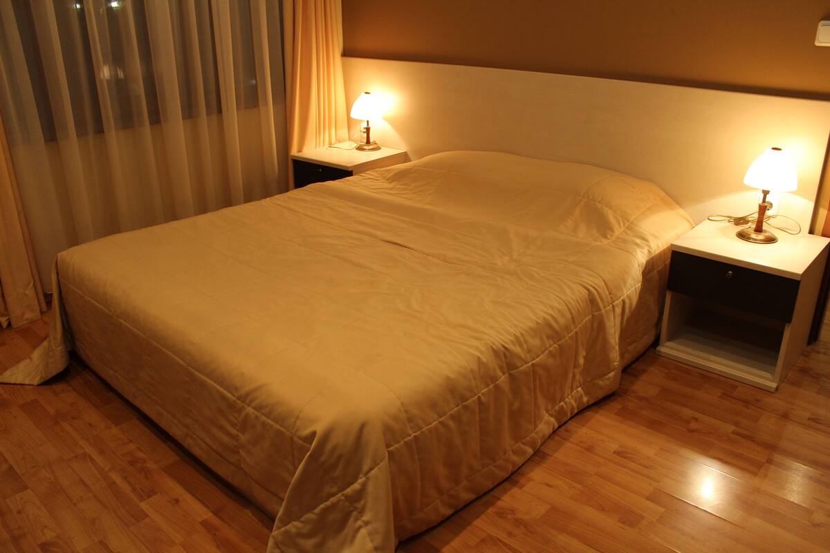 Quarto de hotel com uma cama grande, duas mesas de cabeceira com luminárias, piso de madeira e uma cortina na janela.