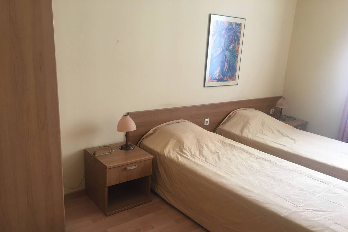 Quarto com duas camas, mesas de cabeceira de madeira, um criado-mudo com abajur e um quadro na parede.
