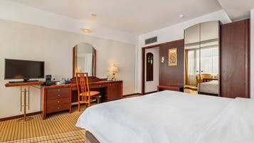 Quarto de hotel com cama, escrivaninha, cadeira, televisão e um espelho grande.