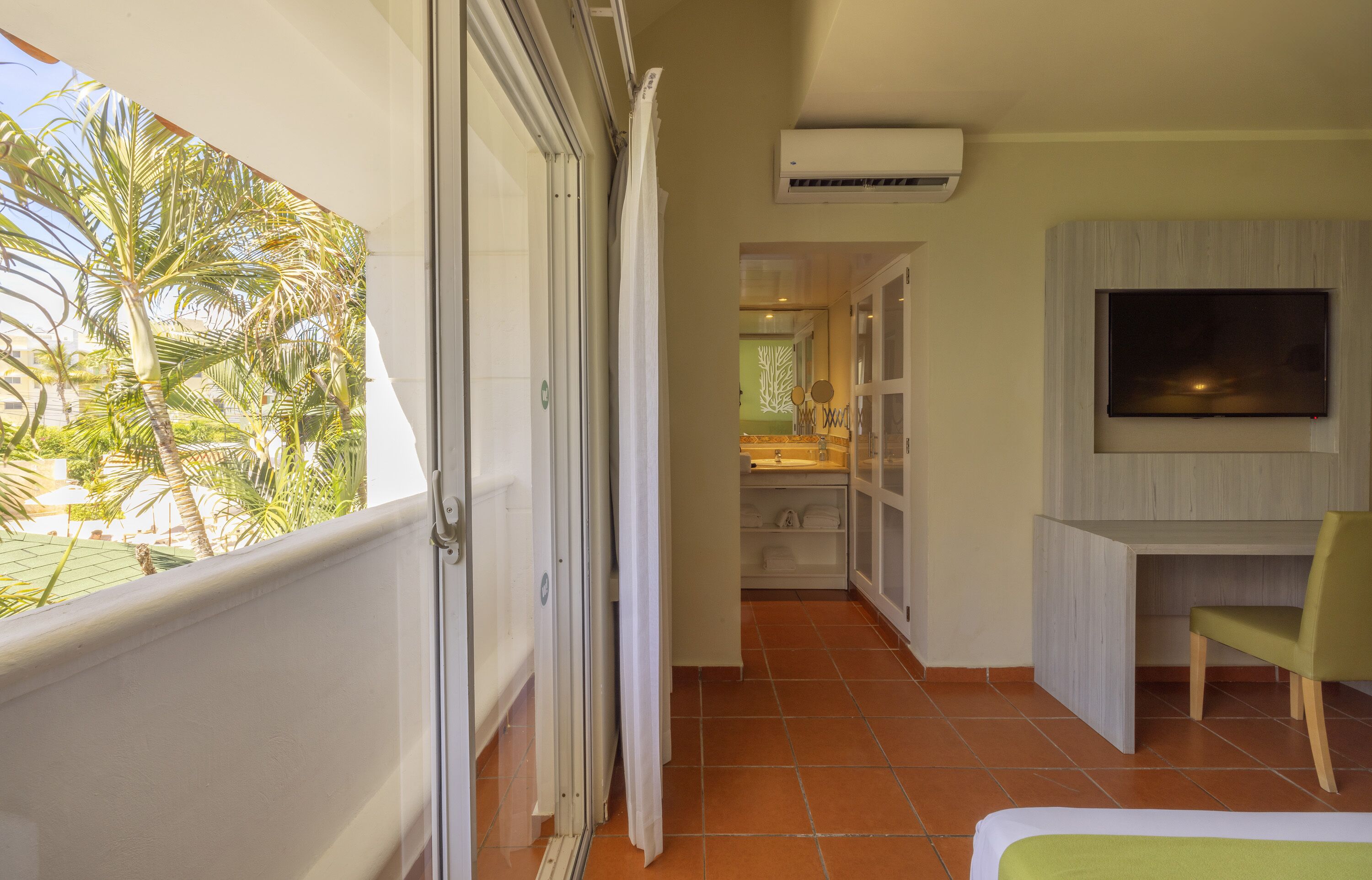 Uma varanda com vista para palmeiras, uma porta de correr, uma área de cozinha com pia e armários, e uma TV fixada na parede.