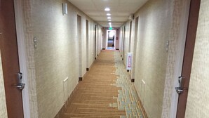 Corredor comprido e estreito de hotel, com carpete estampado, portas em ambos os lados e uma placa na parede direita.