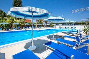 Piscina com cadeiras de sol azuis e guarda-sóis, rodeada por palmeiras e um céu claro.