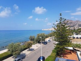 Vista costeira com área de estacionamento, estrada e um edifício com vista para o mar e colinas ao fundo.