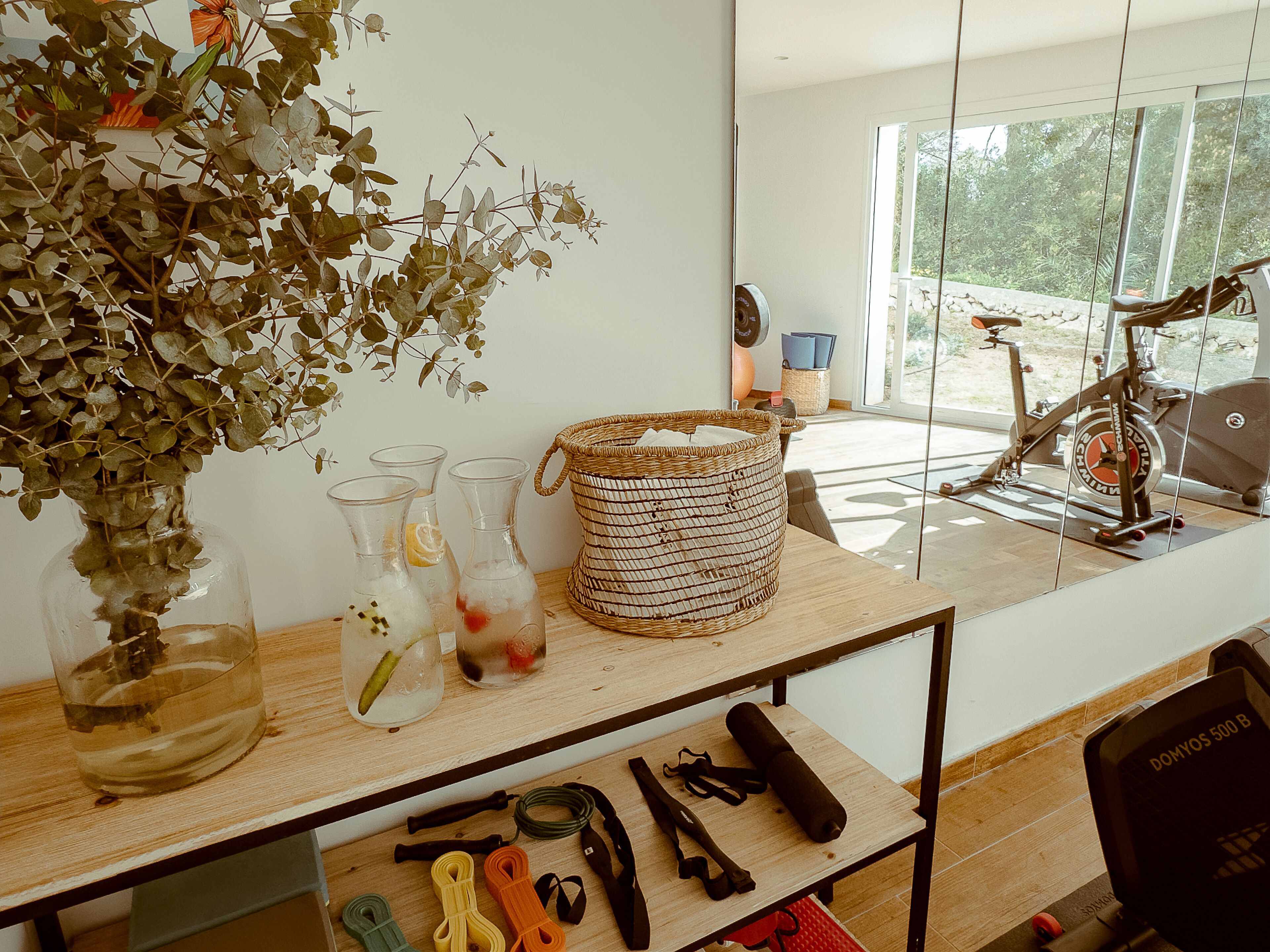 Quarto com uma grande janela, bicicleta ergométrica, cesto de vime e equipamentos de ginástica sobre uma mesa de madeira.