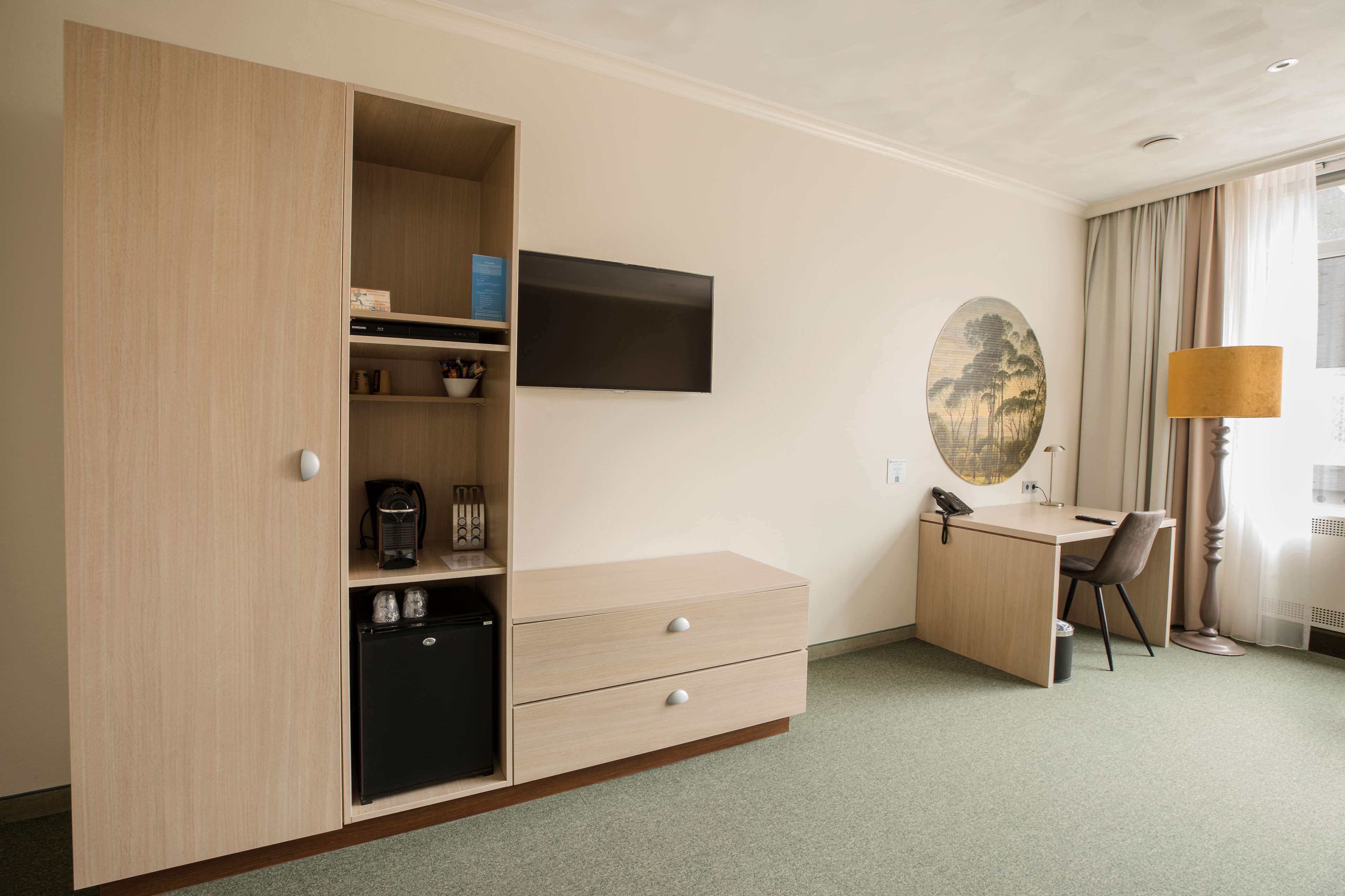 Quarto de hotel com guarda-roupa de madeira, uma escrivaninha com cadeira, TV de tela plana na parede e um pequeno frigorífico.