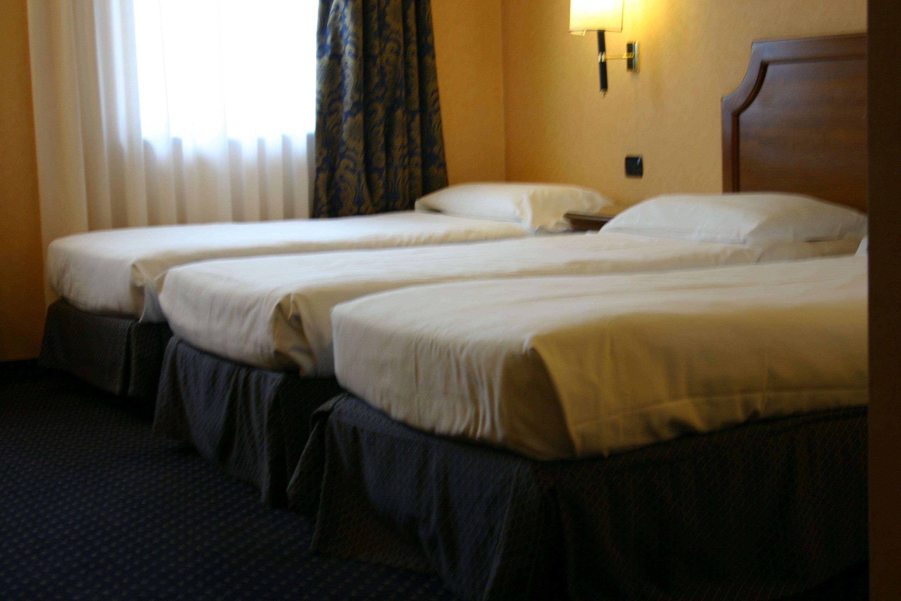 Quarto de hotel com duas camas de solteiro, mesa de cabeceira, luminária e janela com cortinas.