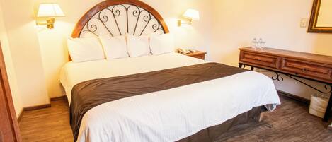 Cama bem arrumada com cabeceira de madeira, mesa de cabeceira com abajur e cômoda de madeira com espelho.