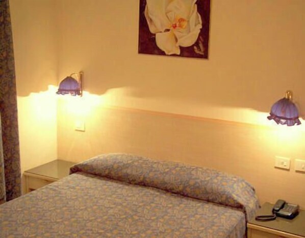 Quarto de hotel com cama, duas luminárias na parede, telefone e um quadro com flores.