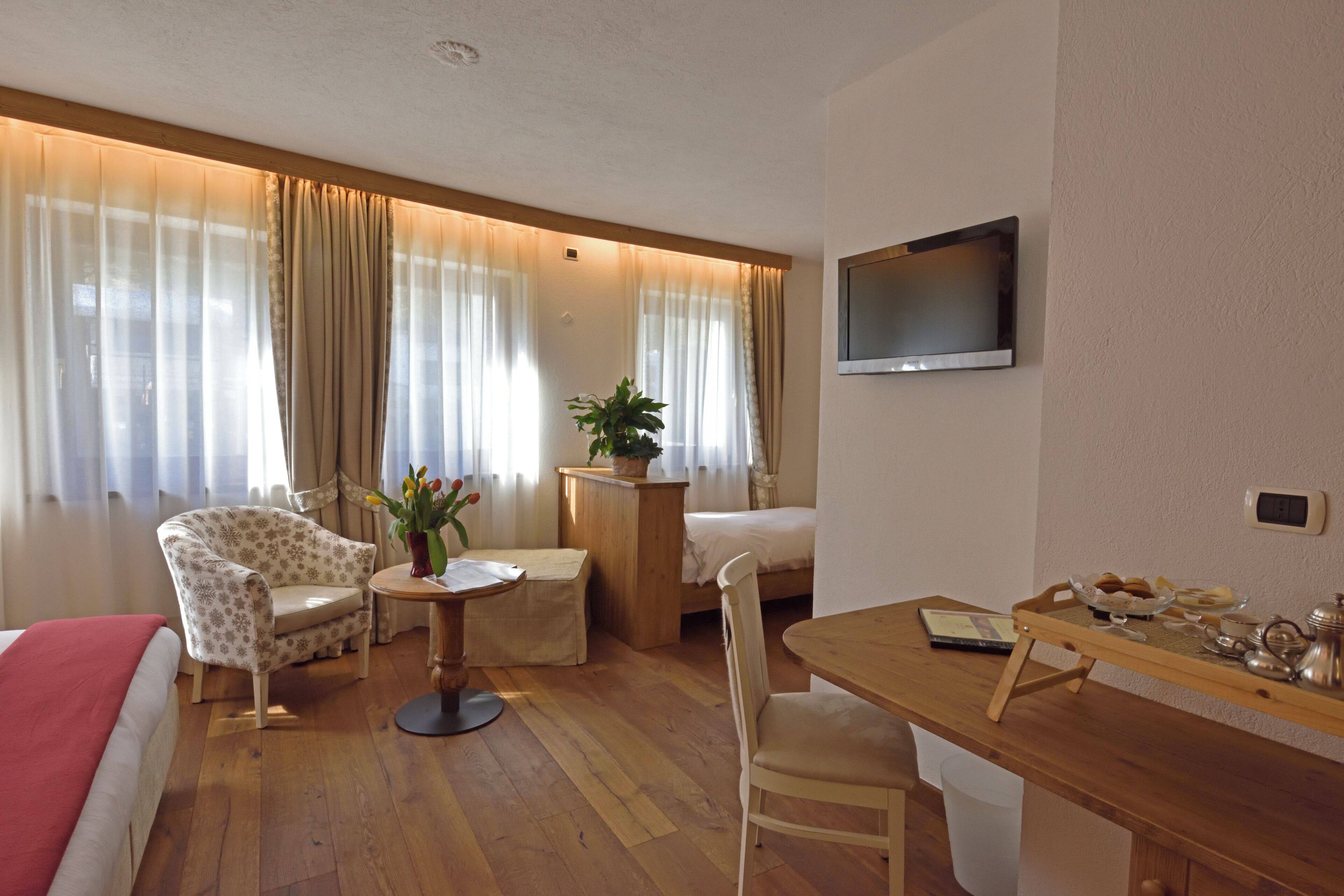 Quarto de hotel com uma cama, uma mesa de jantar de madeira, uma cadeira, uma pequena mesa redonda com uma planta e uma TV na parede.