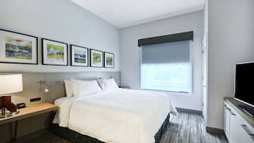 Quarto de hotel com cama, mesa de cabeceira, luminária, obra de arte na parede e janela com persianas.