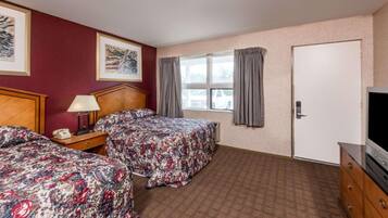 Quarto de hotel com duas camas, televisão, escrivaninha com luminária, porta e janela com cortinas.
