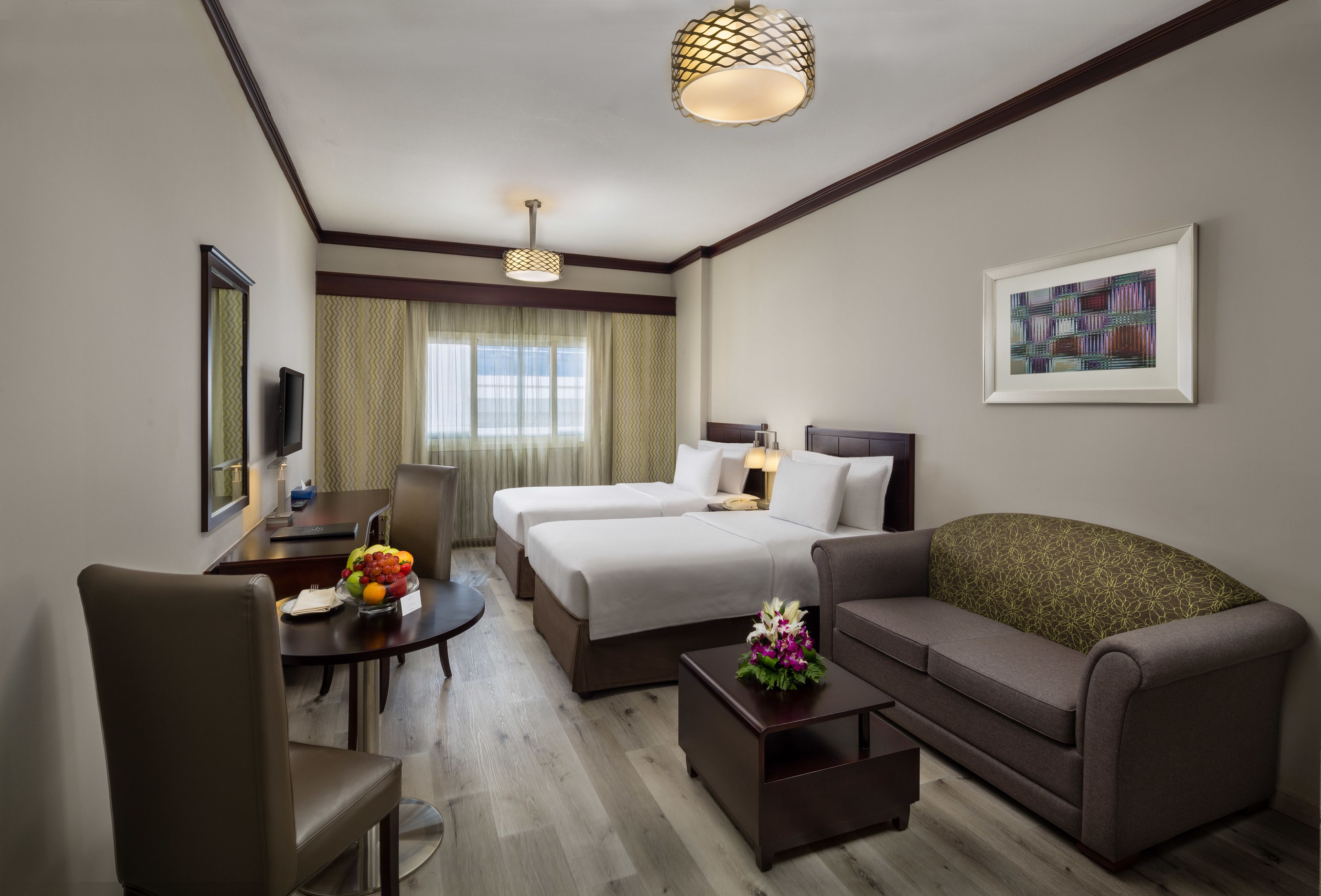 Quarto de hotel com duas camas, um sofá, uma mesa de jantar com frutas, uma televisão e uma obra de arte emoldurada na parede.