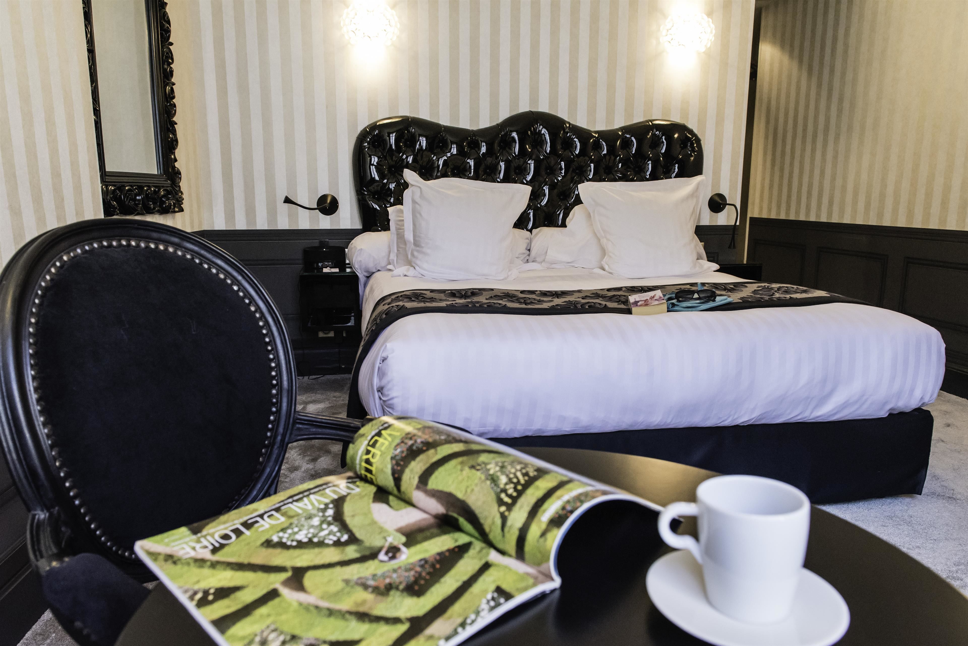 Quarto de hotel com uma cama grande, uma cadeira, uma mesa com uma revista e uma chávena, e um espelho na parede.