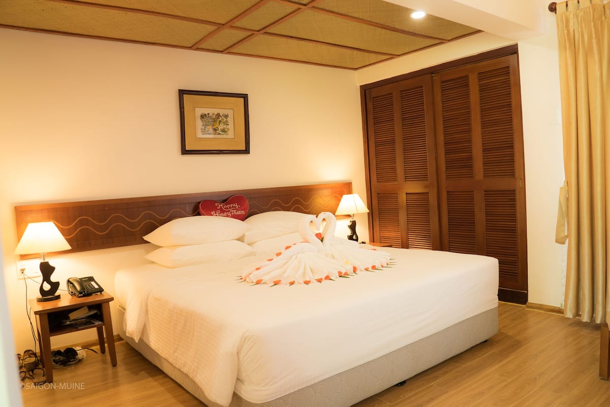 Cama bem arrumada com roupa de cama branca e arranjo decorativo de almofadas em formato de cisne. Tem uma mesa de cabeceira com luminária e telefone. Um quadro emoldurado está pendurado na parede acima da cama.