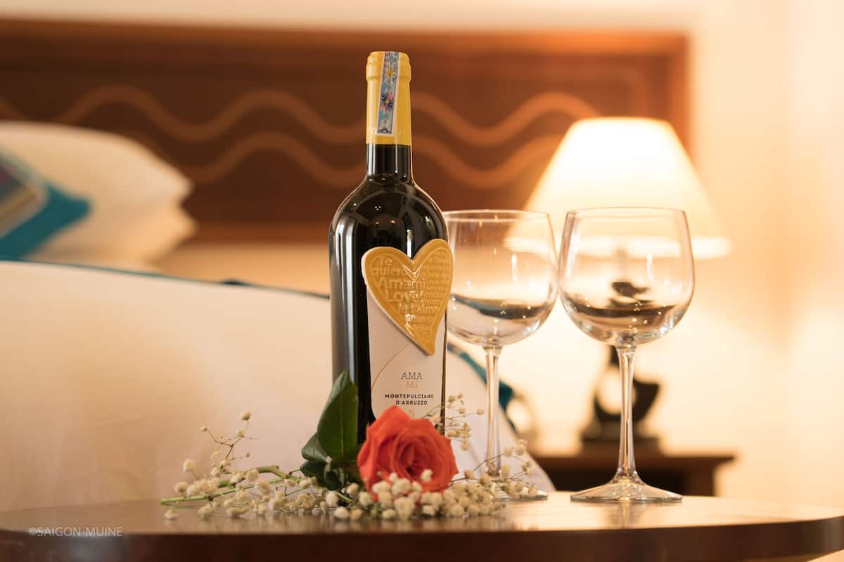 Uma garrafa de vinho Montemaggi, uma rosa e dois copos de vinho sobre uma mesa em quarto de hotel.