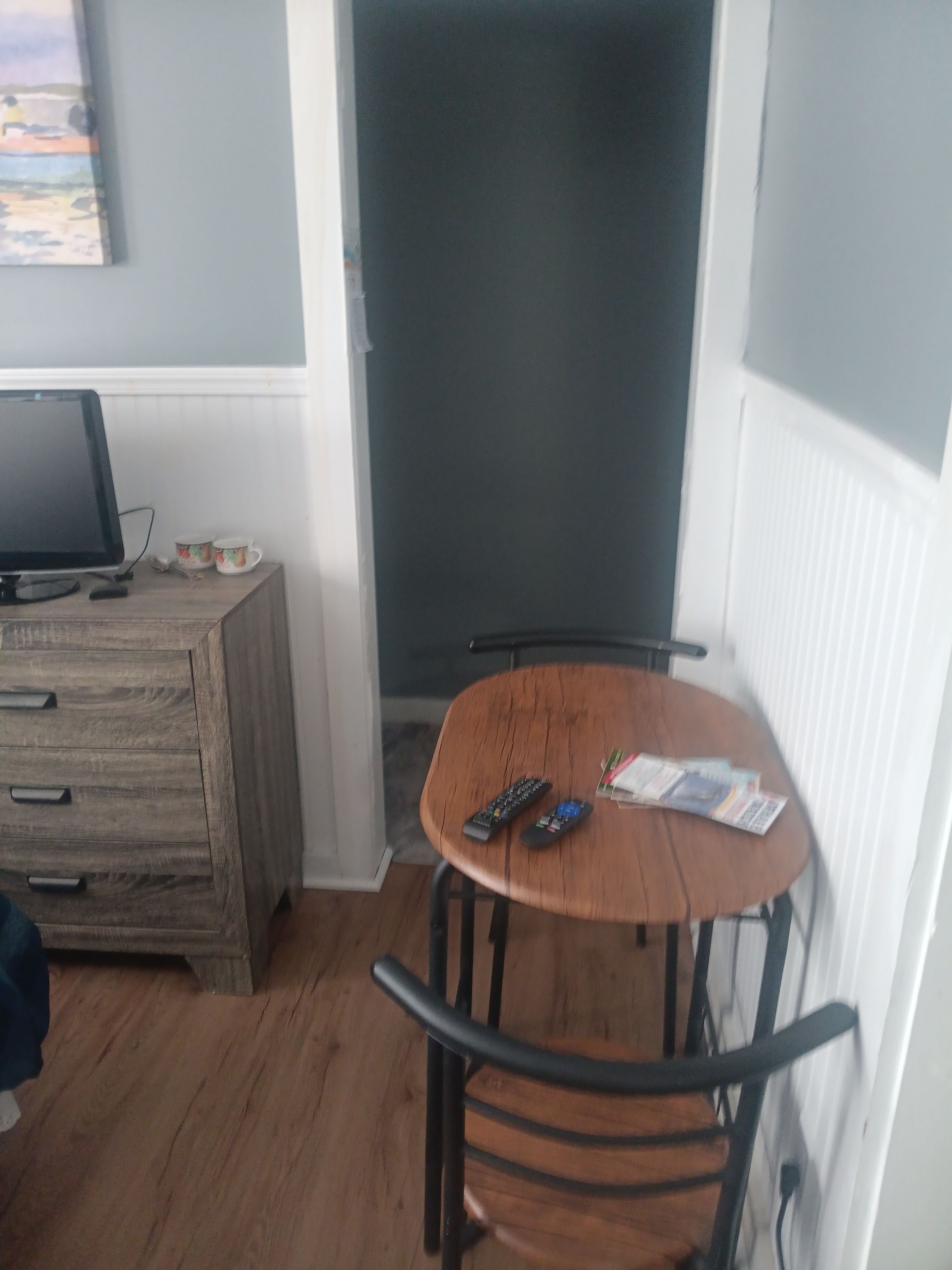 Quarto com cômoda de madeira, uma mesinha com controles remotos e papéis, e um quadro na parede.