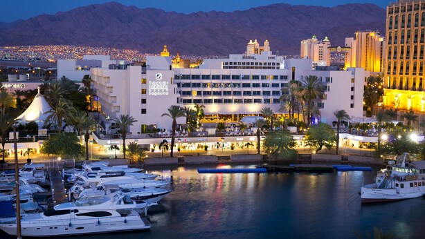 Uma marina com barcos atracados, um hotel com vários andares e um cenário montanhoso ao fundo.