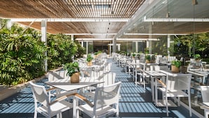 Um café ao ar livre com mesas e cadeiras brancas, rodeado por vegetação exuberante e uma pérgola de madeira.
