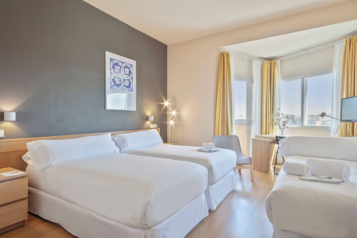 Quarto de hotel com duas camas, uma escrivaninha, uma cadeira, uma televisão e uma janela com cortinas.