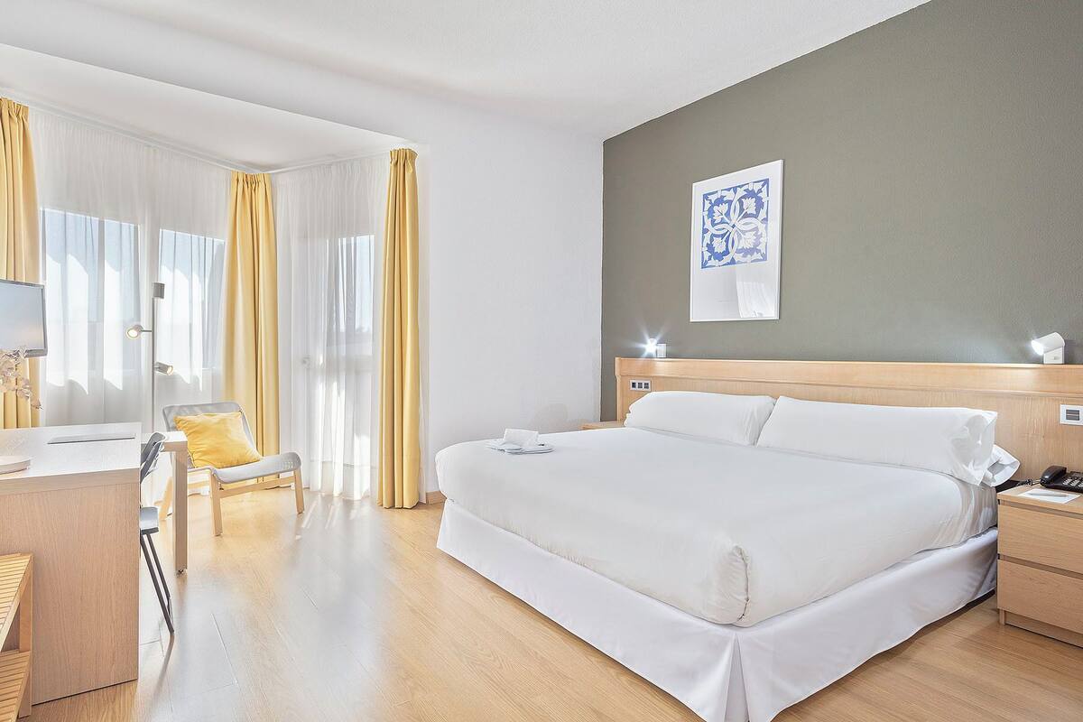 Quarto de hotel com cama, mesa de cabeceira, escrivaninha, cadeira e uma janela grande com cortinas.