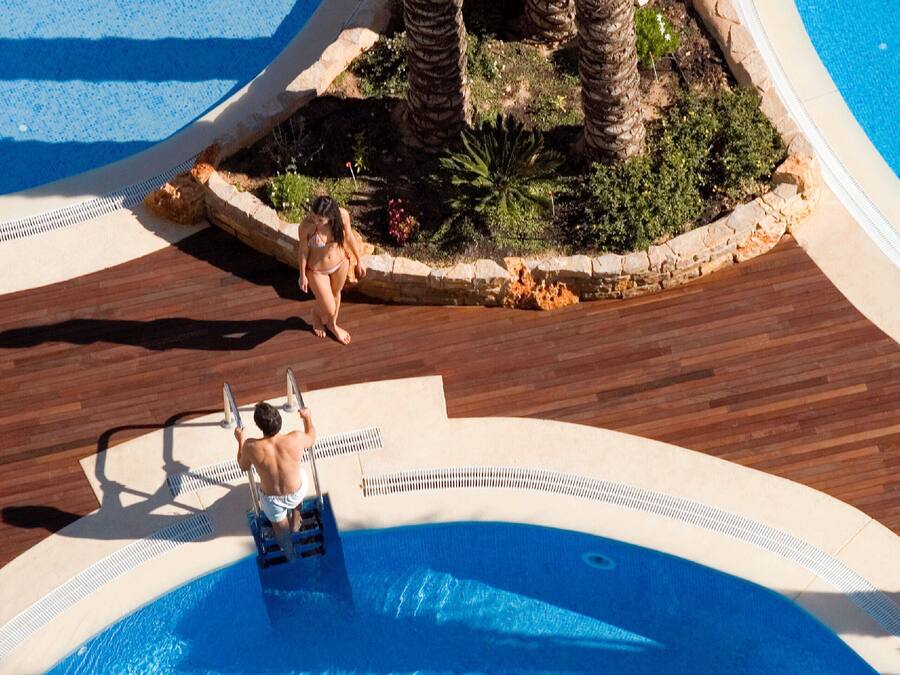 Uma área de piscina com deck de madeira, plataforma de mergulho e uma pequena ilha com palmeiras e plantas.
