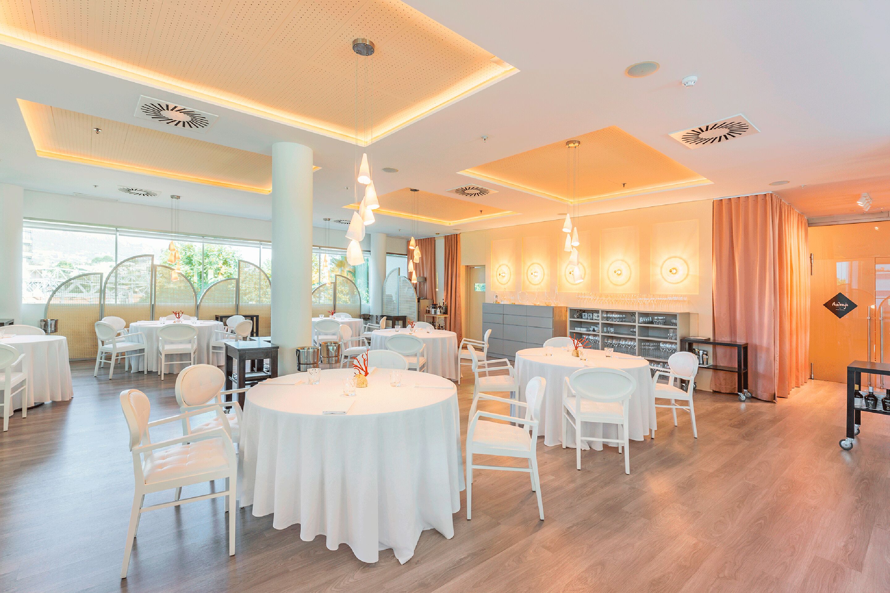 Uma ampla sala de jantar de hotel com mesas redondas e cadeiras brancas, janelas grandes e um design moderno no teto.