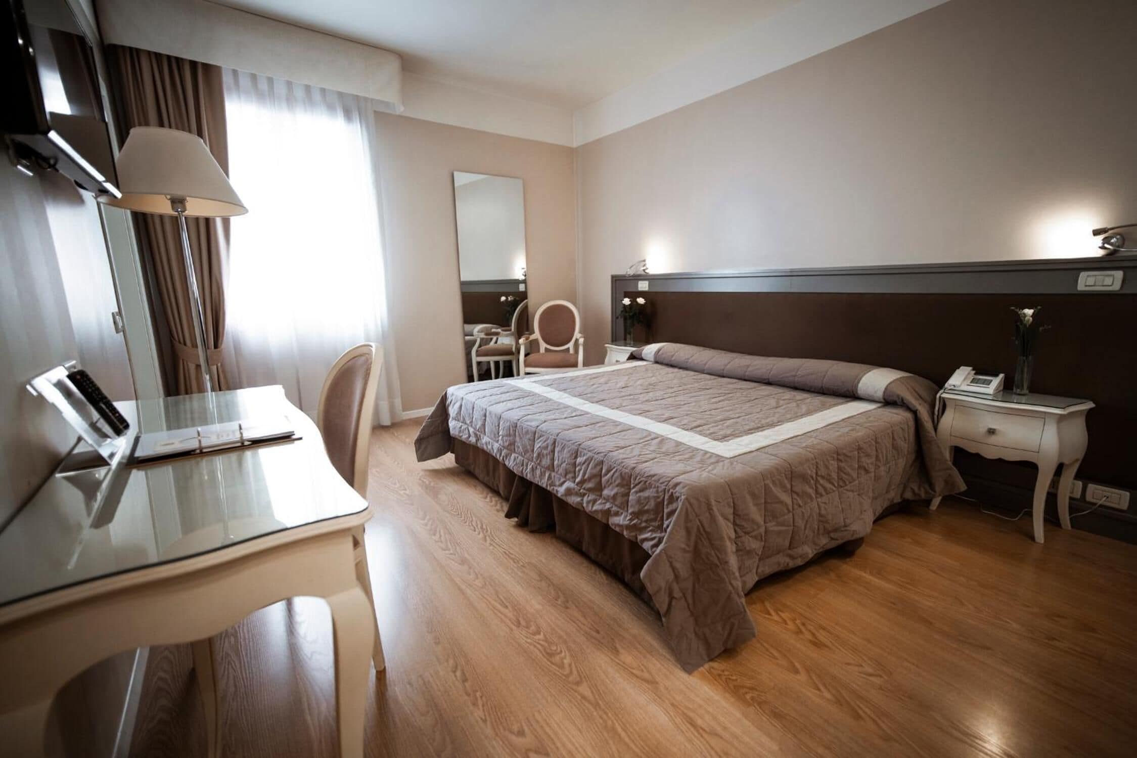 Quarto de hotel com cama, escrivaninha com luminária, cadeira, mesa de cabeceira com telefone e espelho.