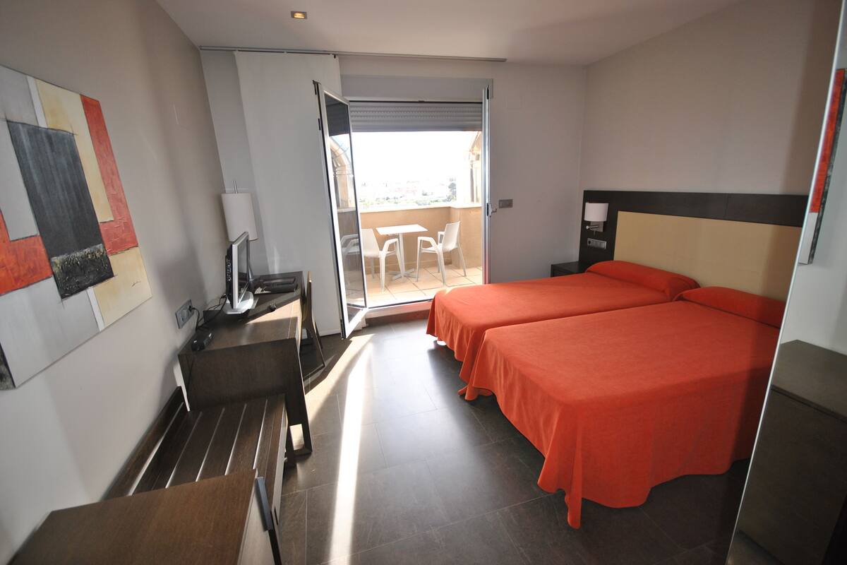 Quarto de hotel com duas camas, uma escrivaninha, uma cadeira, uma TV e uma varanda com cadeiras.