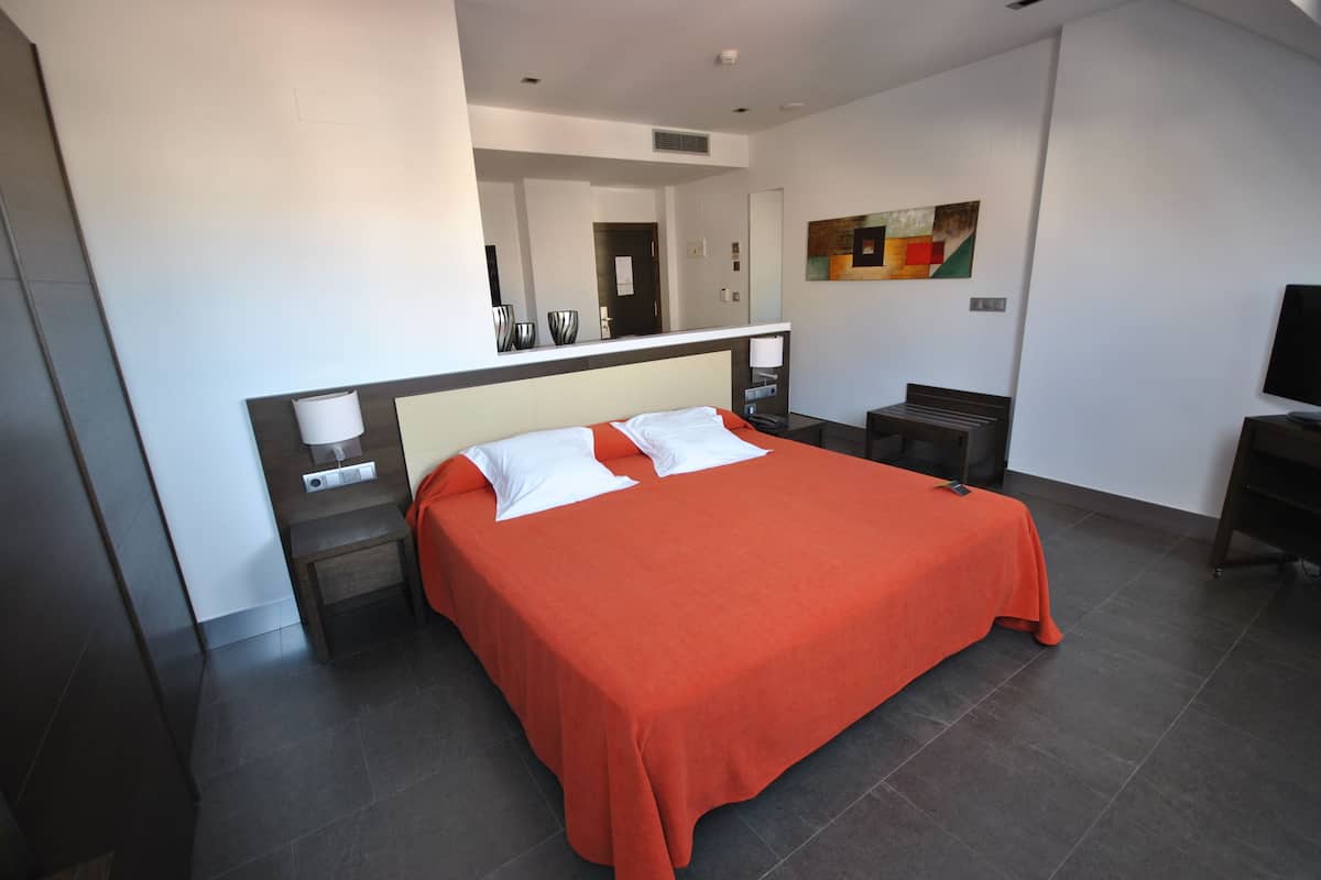 Quarto de hotel moderno com uma cama grande, mesa de cabeceira, escrivaninha, cadeira, televisão e um quadro na parede.