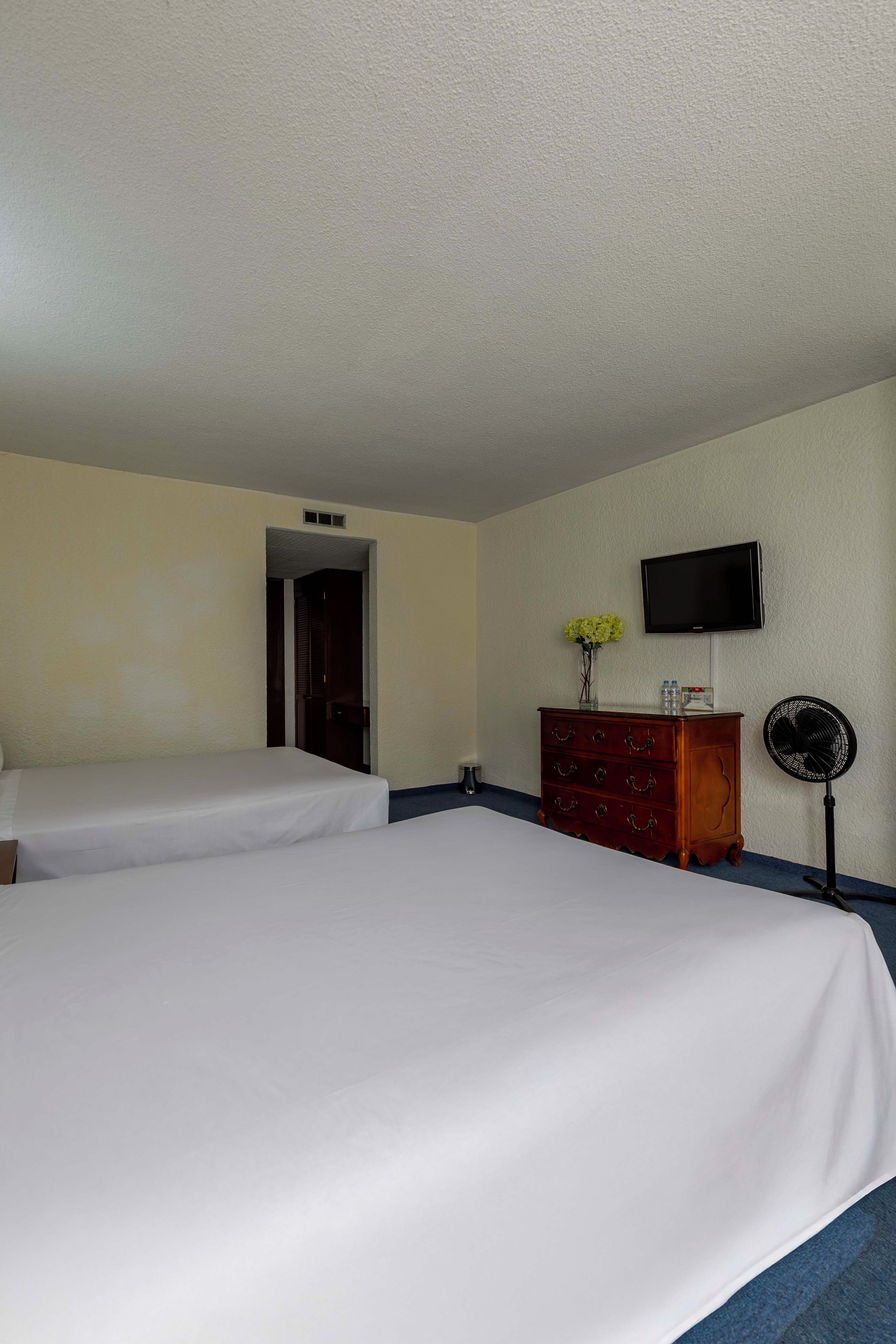 Quarto de hotel com uma cama grande, cômoda, televisão e ventilador.