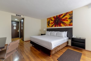 Quarto de hotel com uma cama grande, uma mesinha de cabeceira, uma escrivaninha, uma cadeira, um quadro na parede e piso de madeira.