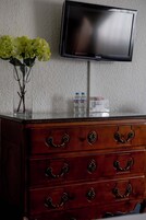 Uma cômoda de madeira com puxadores decorados, um vaso com flores, duas garrafas de água e uma televisão fixada em uma parede texturizada.