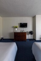 Quarto de hotel com duas camas, cômoda, TV de tela plana, ventilador e um vaso com flores.