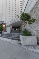 Entrada de um hotel com design moderno, apresentando uma escadaria que leva à entrada, uma grande placa acima da porta e uma área ajardinada com plantas.