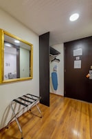 A room with a wooden floor, a mirror with a gold frame, a black ironing board, and a door with a hanging paper.