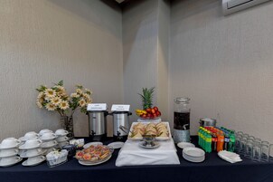 Disposição de buffet com dispensadores de café e chá, uma cesta de frutas, sanduíches e diversas bebidas.